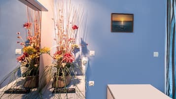 Casa de banho com parede azul, uma fotografia emoldurada de um pôr do sol e dois arranjos florais numa estante.