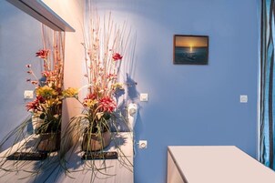 Casa de banho com parede azul, uma fotografia emoldurada de um pôr do sol e dois arranjos florais numa estante.