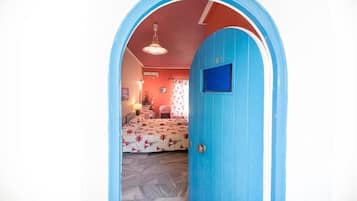 Um quarto com porta azul, cama com roupa de cama florida, parede vermelha e luminária pendente.