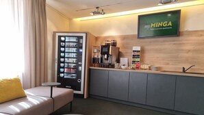 Um lobby moderno de hotel com um sofá, uma máquina de café, uma máquina de venda automática e um balcão com uma placa indicando "my MINGA Hotel & serviced Apartments".