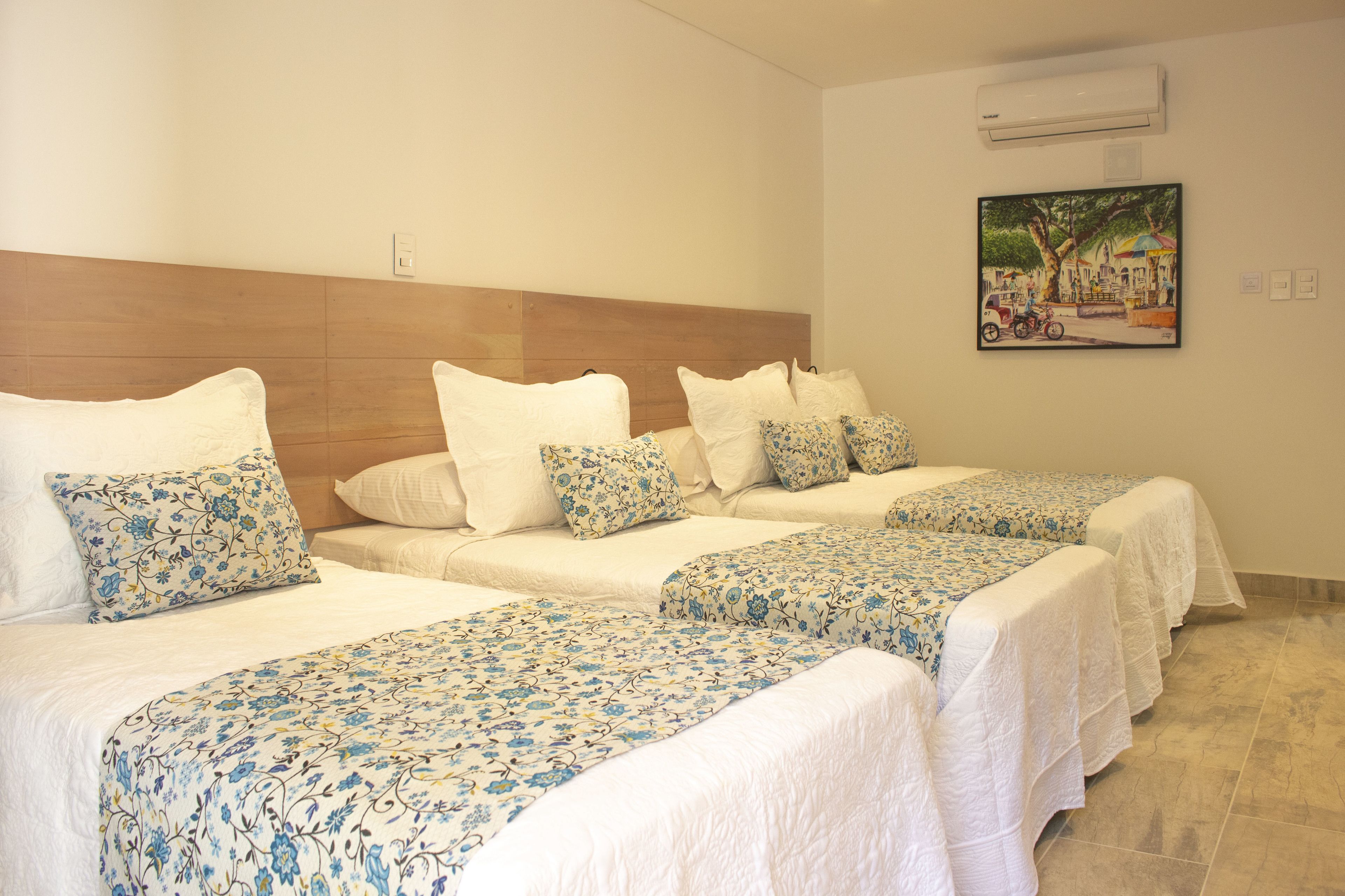 Quarto de hotel com três camas, cada uma com roupa de cama branca e estampas florais azuis. Há um quadro na parede retratando uma cena de rua.