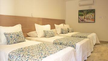 Quarto de hotel com três camas, cada uma com roupa de cama branca e estampas florais azuis. Há um quadro na parede retratando uma cena de rua.