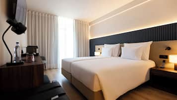 Quarto de hotel moderno com uma cama grande, mesas de cabeceira, escrivaninha com abajur e televisão na parede.