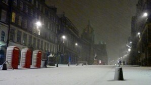 Rua coberta de neve à noite, com edifícios e postes de luz.