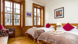 Quarto de hotel com duas camas, piso de madeira, janelas amplas, uma cadeira e obras de arte nas paredes.