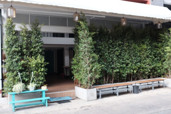 Área externa com mesas e bancos, plantas em vasos, galeria coberta e entrada de um edifício.
