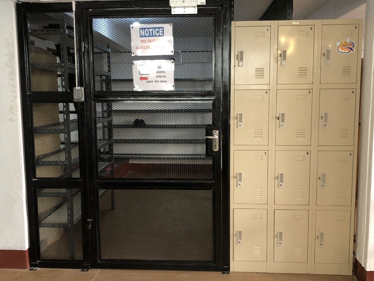 Uma porta de segurança com uma placa de aviso, uma porta mosquiteiro e um conjunto de armários bege com portas numeradas.