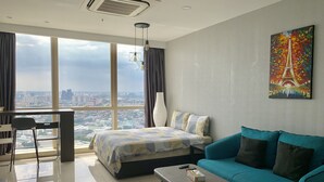 Quarto de hotel moderno com uma cama grande, um sofá azul, uma mesa pequena e vista da cidade através de janelas amplas.