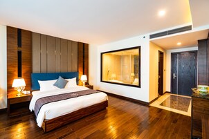 Quarto de hotel moderno com uma cama grande, piso de madeira, cabeceira azul e uma janela ampla.