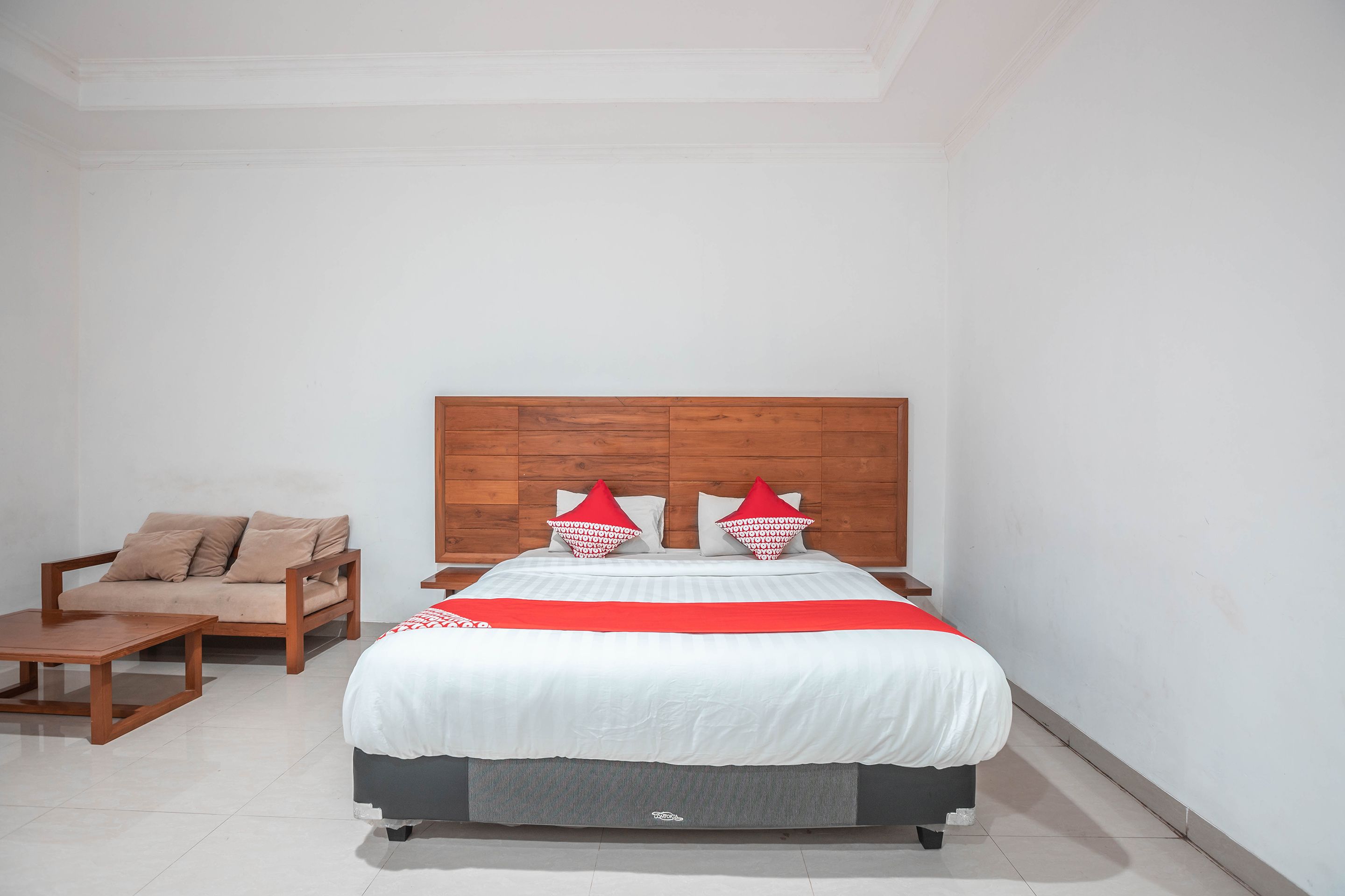 Quarto com cabeceira de madeira, cama com roupa de cama branca e almofadas vermelhas, uma cadeira de madeira e uma mesa pequena.