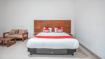 Quarto com cabeceira de madeira, cama com roupa de cama branca e almofadas vermelhas, uma cadeira de madeira e uma mesa pequena.