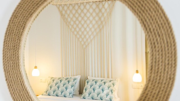 Espelho redondo com moldura de fibra natural refletindo uma cama com duas almofadas, luminária pendente e luz fixa na parede.
