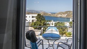 Varanda com mesa e cadeiras, um guarda-sol azul e vista para paisagem costeira com edifícios e colinas.