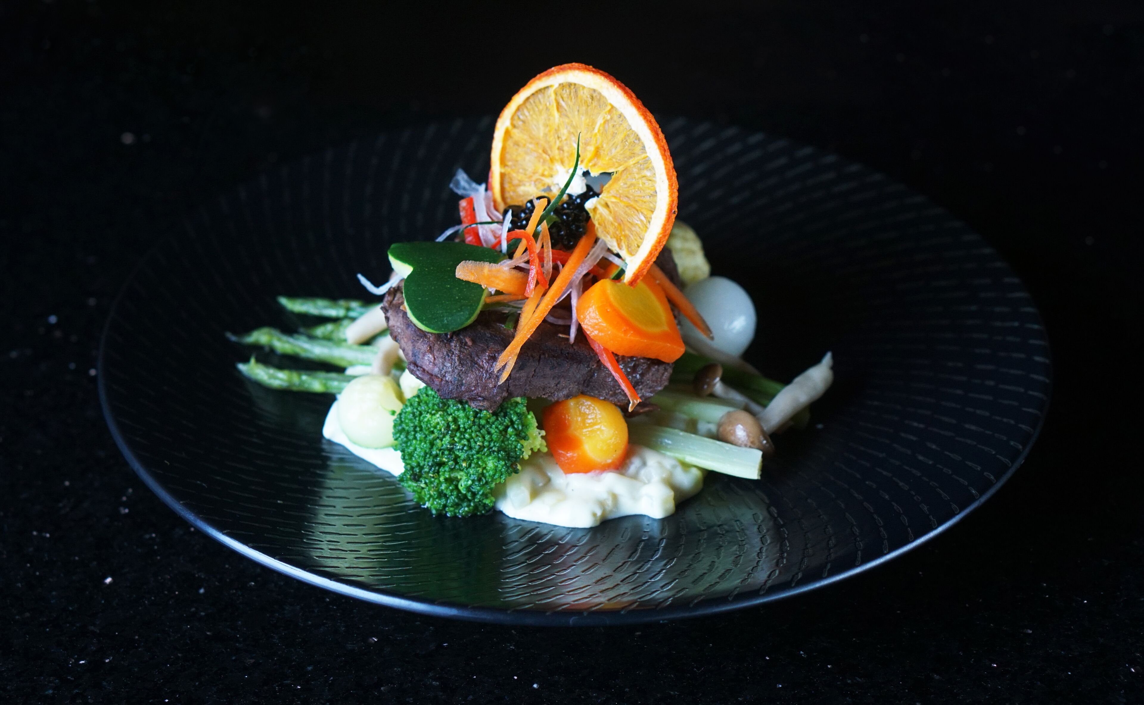 Um prato negro com diversos alimentos, incluindo um pedaço de carne, legumes e uma fatia de laranja.