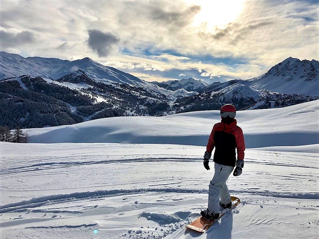 Um snowboarder numa montanha nevada, com vista para picos e árvores cobertas de neve ao fundo.