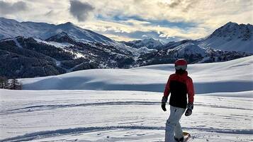 Um snowboarder numa montanha nevada, com vista para picos e árvores cobertas de neve ao fundo.