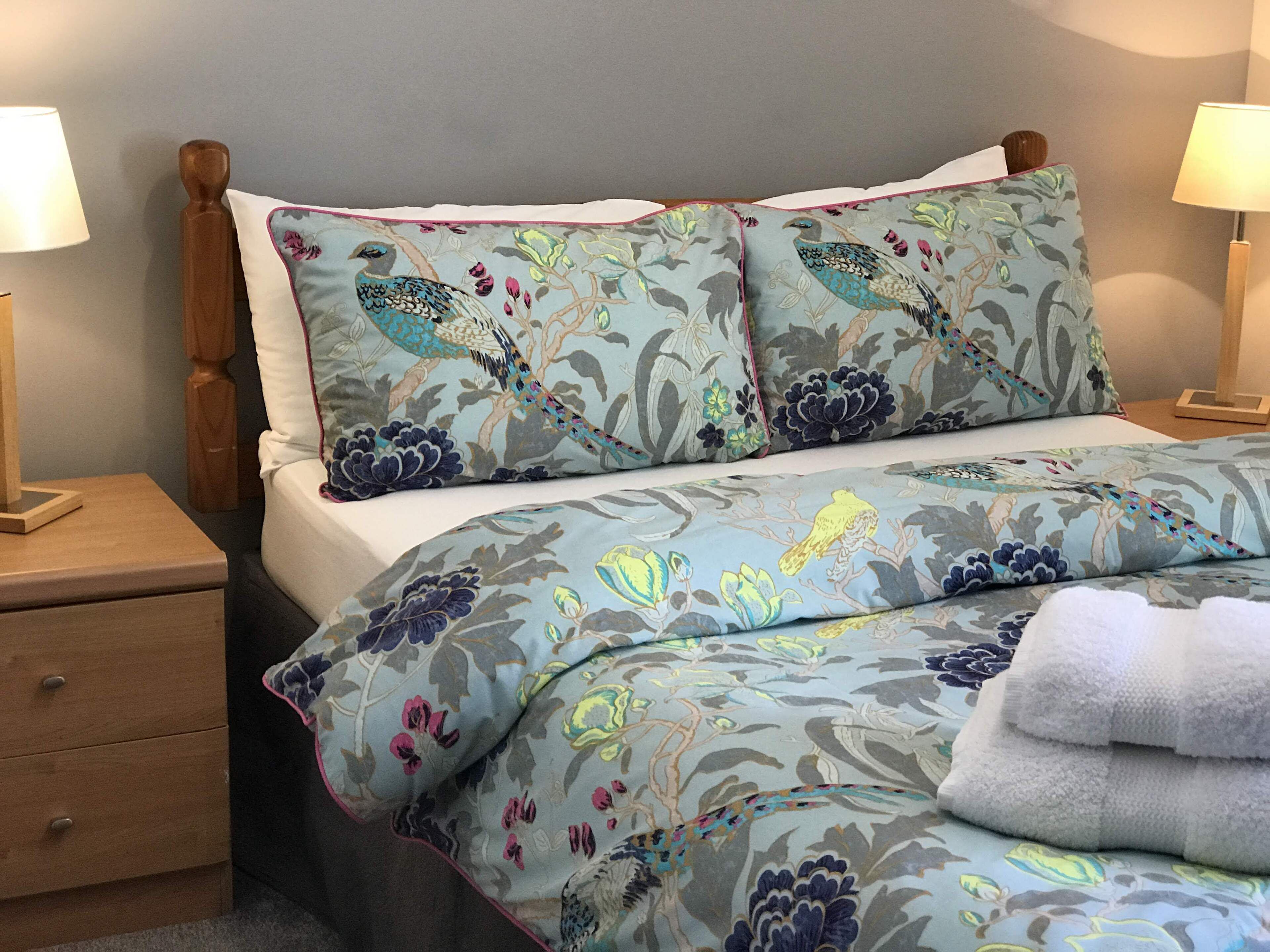 Uma cama bem arrumada com roupa de cama floral, duas luminárias de cabeceira e uma mesinha de cabeceira de madeira com gaveta.