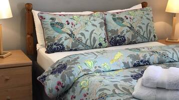 Uma cama bem arrumada com roupa de cama floral, duas luminárias de cabeceira e uma mesinha de cabeceira de madeira com gaveta.
