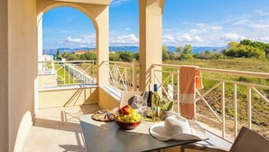 Uma varanda com uma mesa posta para refeição, uma tigela de frutas, garrafas de vinho e um chapéu.