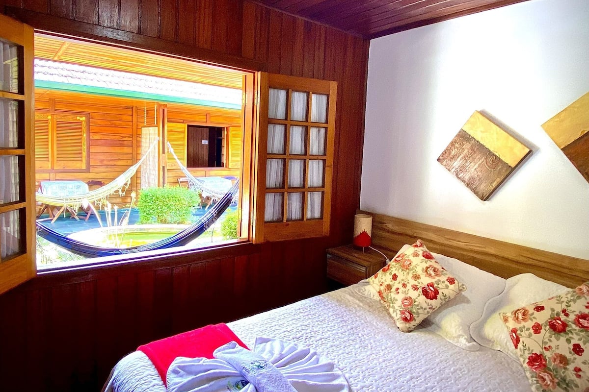 Quarto de cabana de madeira com cama, rede e vista para um edifício de madeira lá fora.