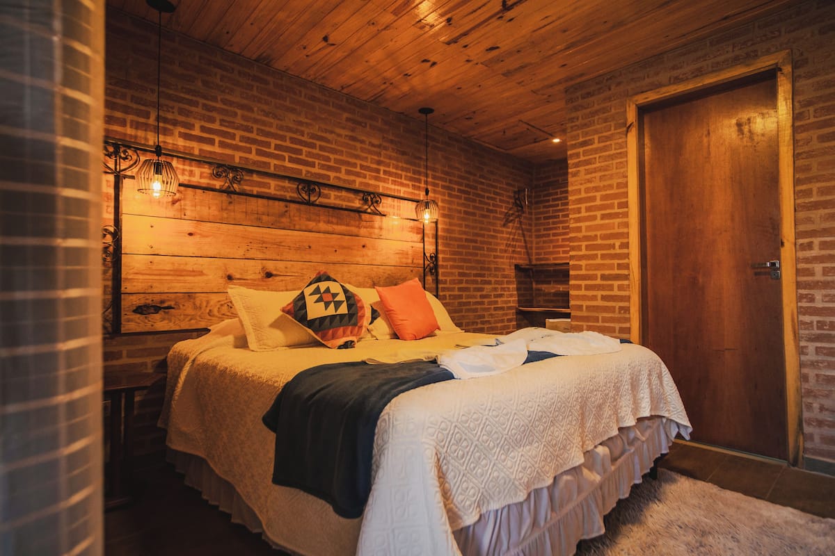 Quarto com parede de tijolos, teto de madeira e cama com várias almofadas e um cobertor.