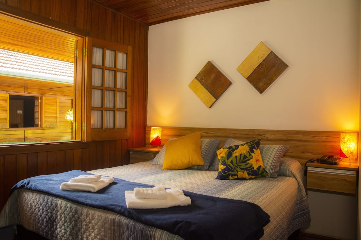 Quarto de hotel com uma cama, duas pinturas penduradas na parede, uma janela com persianas, um abajur e um telefone na mesa de cabeceira.
