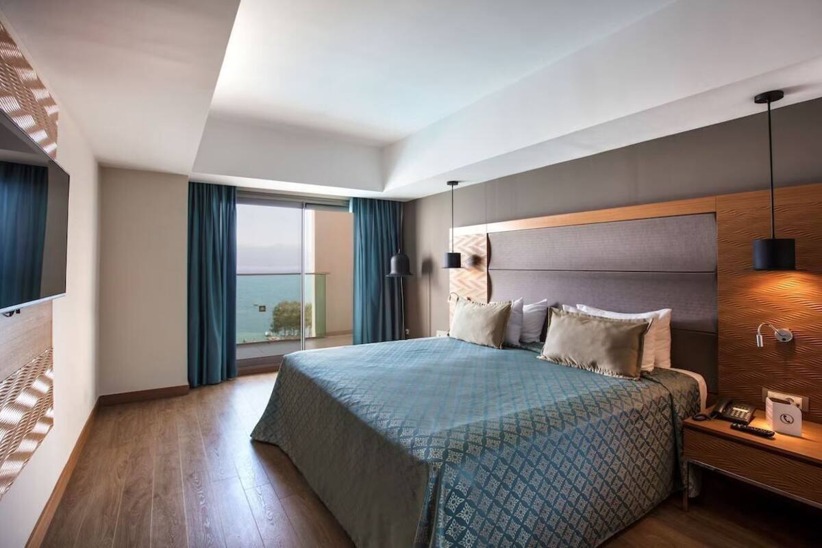 Quarto de hotel moderno com uma cama grande, televisão, cabeceira de madeira e varanda com vista.