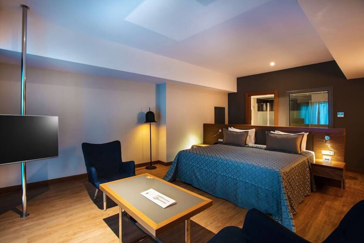 Quarto de hotel com cama, secretária, cadeira, televisão, mesa de cabeceira e abajur.