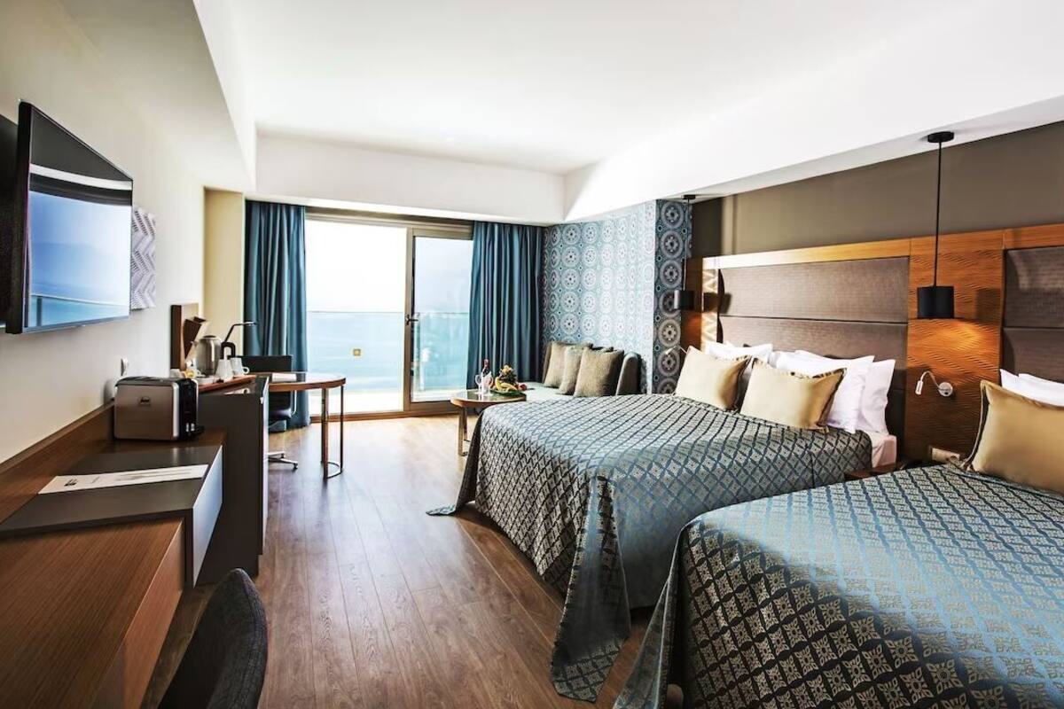 Quarto de hotel com duas camas, uma mesa, uma TV e uma varanda com vista.