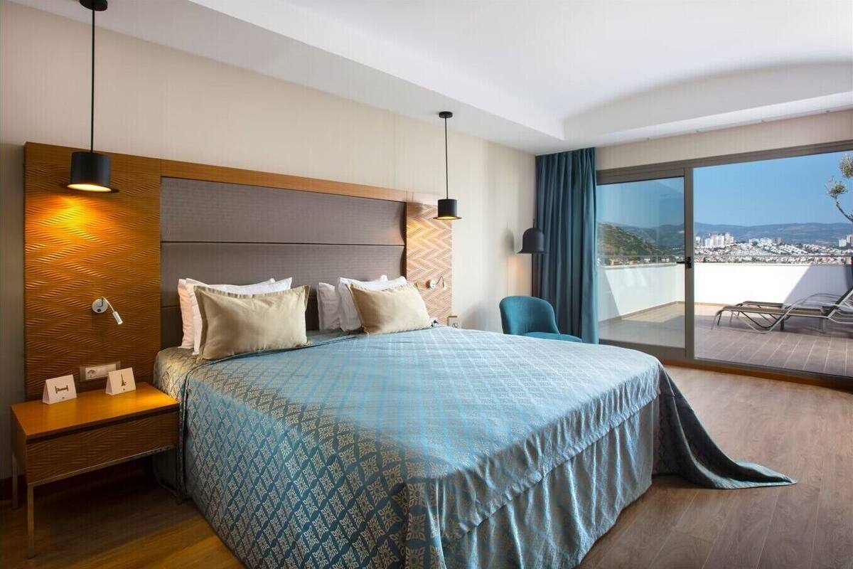 Quarto de hotel com cama grande, mesa de cabeceira, escrivaninha, cadeira e varanda com vista.