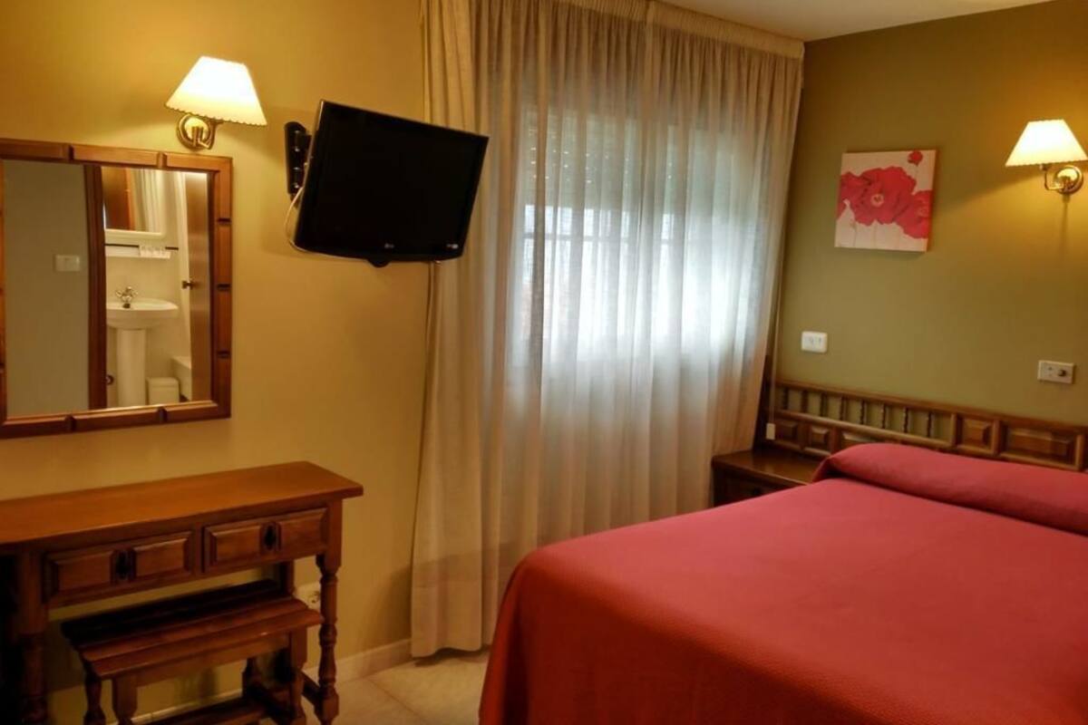 Quarto de hotel com uma cama, televisão na parede, cómoda de madeira, espelho, janela com cortinas e um quadro na parede.