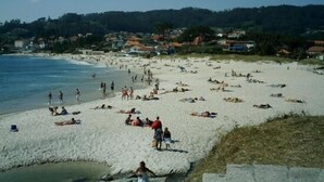 Uma praia com pessoas tomando banho de sol e nadando, cercada por casas e vegetação.