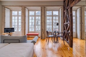 Quarto de hotel com cama, escrivaninha com cadeira, televisão e janelas amplas com vista para edifícios.