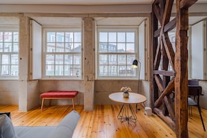 Quarto com grandes janelas, piso de madeira, mesa redonda com um vaso, um banco e estrutura de vigas de madeira.