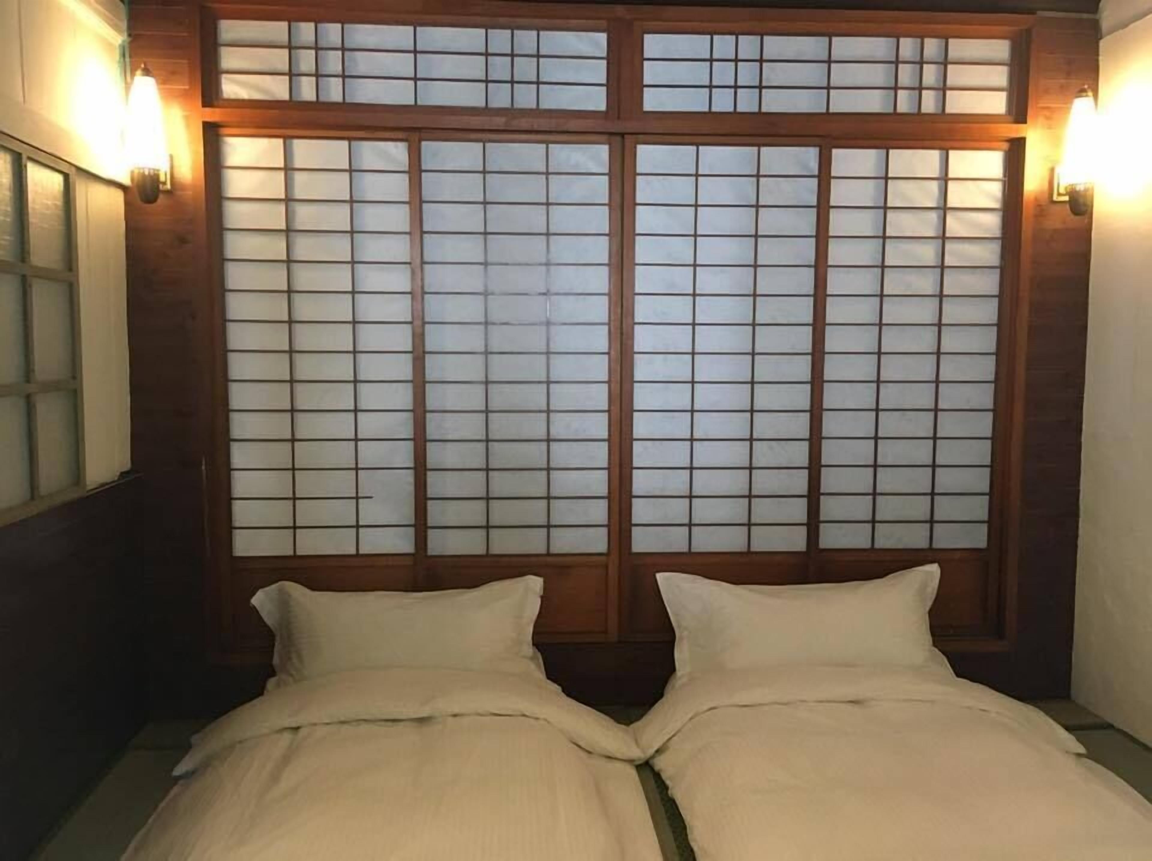 Quarto com porta tradicional de shoji japonesa, duas camas com roupa de cama branca e arandelas na parede que fornecem iluminação.
