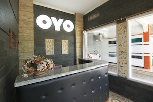 Balcão de recepção com uma grande placa da marca "OYO", um vaso de flores e vista para um trem através de uma porta de vidro.
