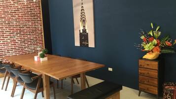 Uma área de refeições com uma mesa e cadeiras de madeira, um banco, um quadro pendurado na parede retratando um edifício, uma pequena planta e um vaso decorativo com flores.