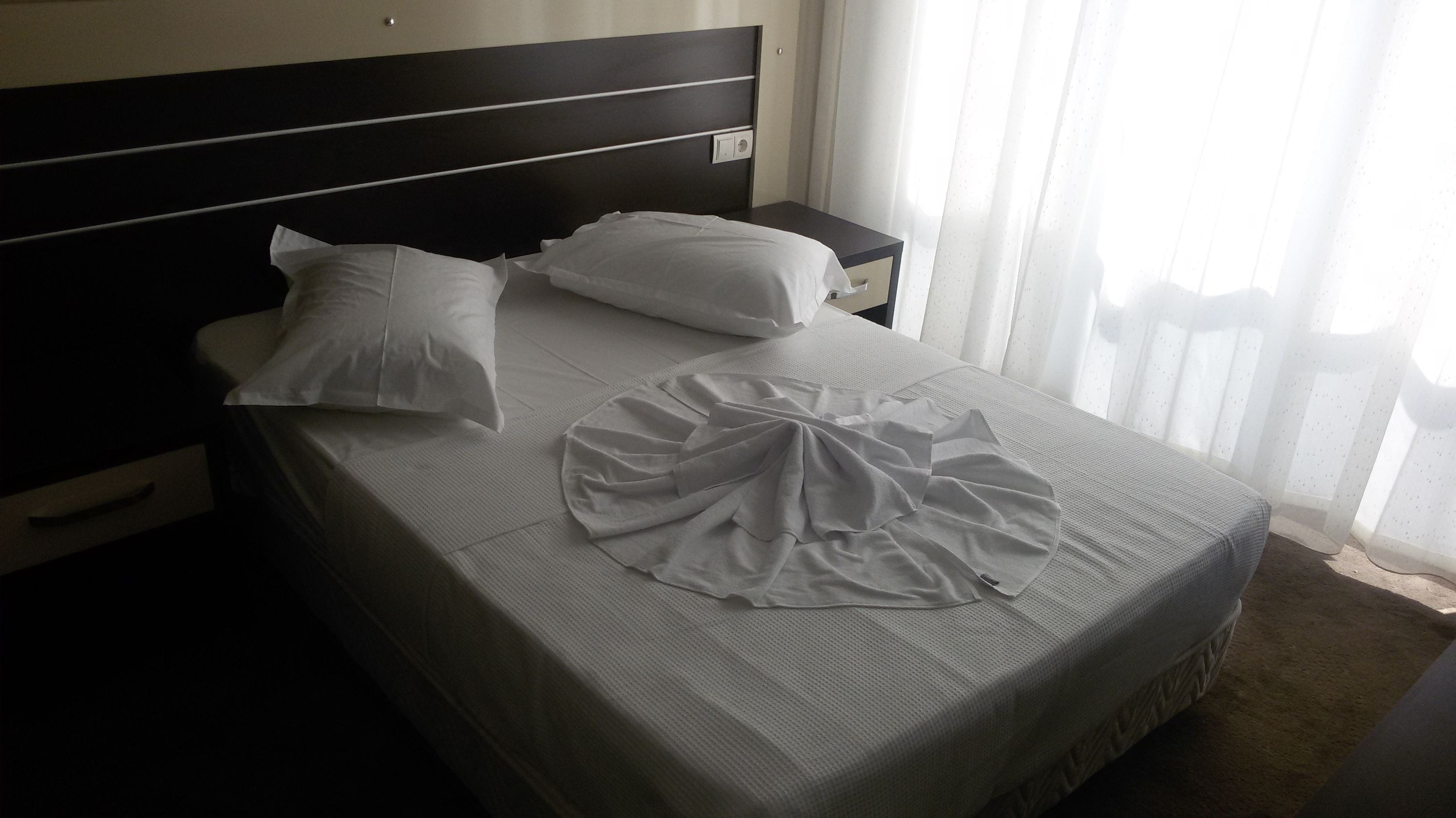 Uma cama bem arrumada com roupa de cama e almofadas brancas, uma mesa de cabeceira e uma janela com cortinas leves.