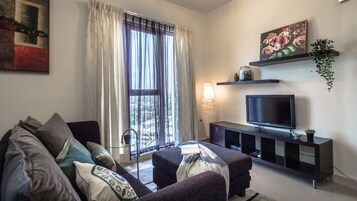 Uma sala de estar com sofá, televisão em um móvel, janela com cortinas e um quadro na parede.