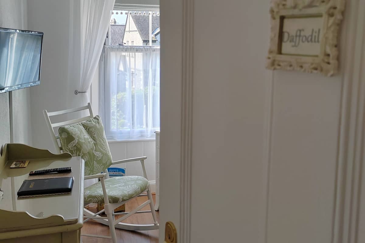 Quarto com uma cadeira de balanço, uma mesinha com um controle remoto e um livro, uma janela com cortinas e uma porta com a placa 'Daffodil'.