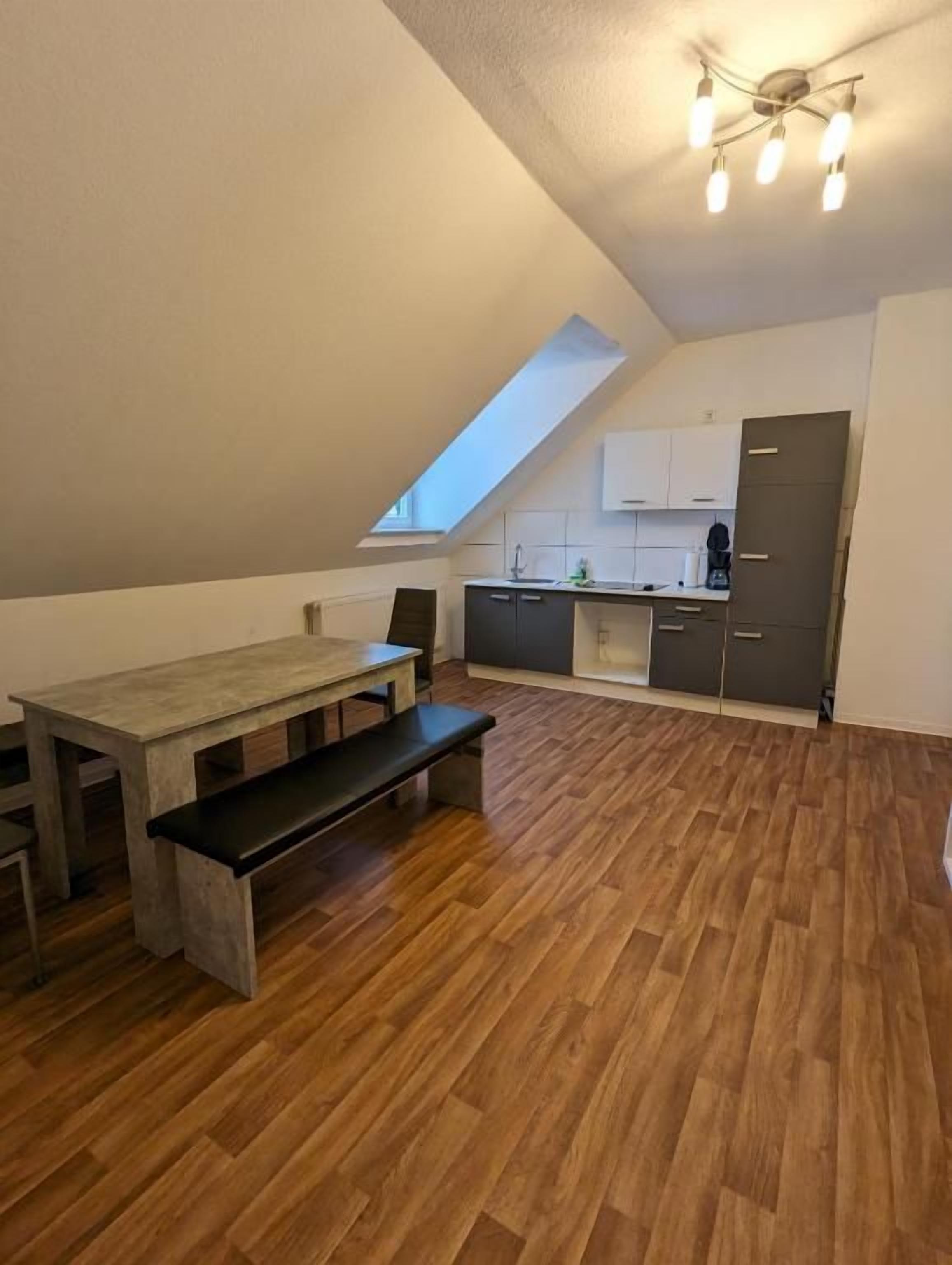 Um apartamento em estilo loft, com teto inclinado, área de jantar com banco, cozinha compacta e uma janela grande.