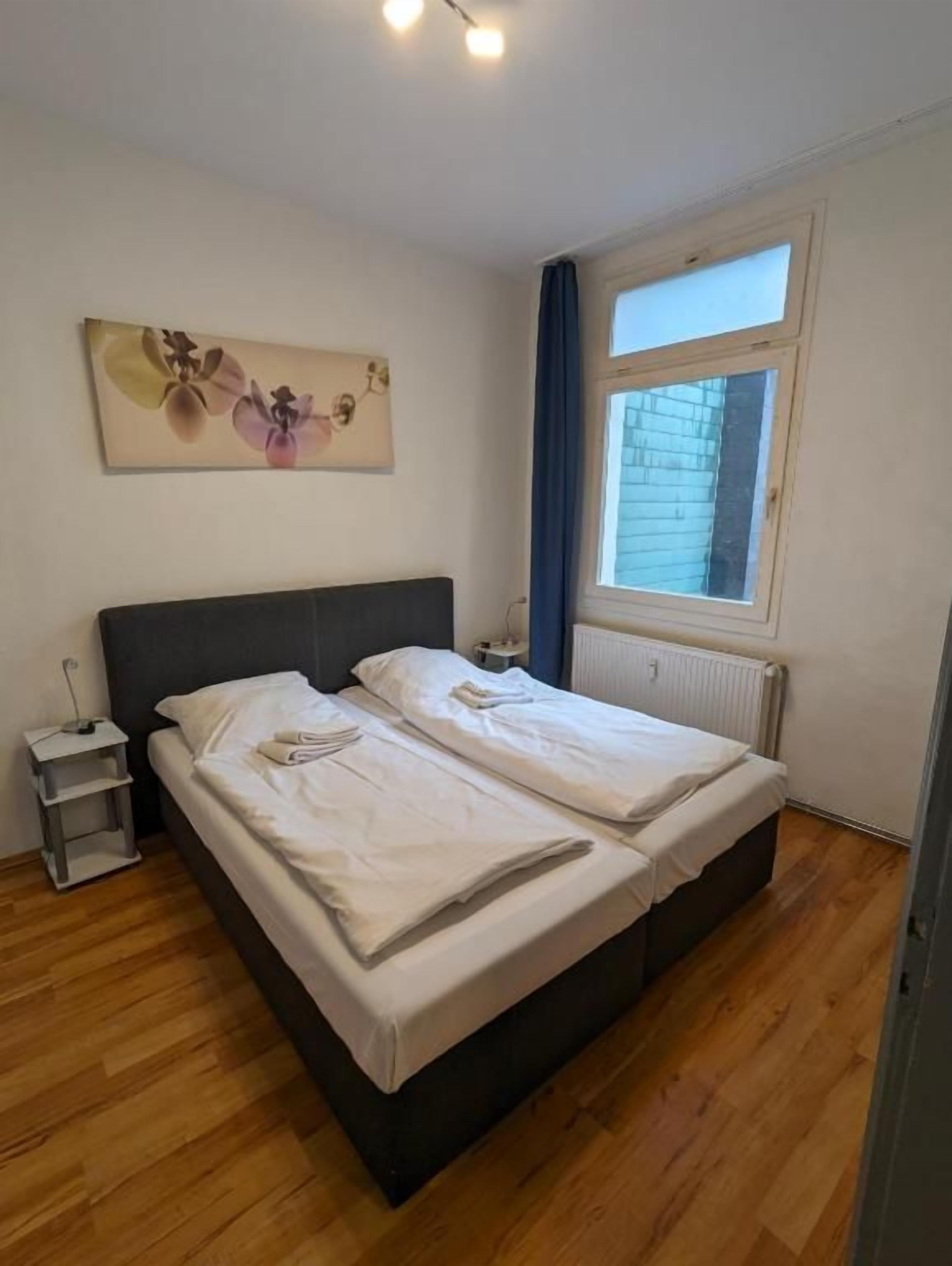 Uma cama de casal com lençóis brancos, uma mesinha de cabeceira, uma obra de arte na parede, uma janela com cortinas e um radiador.