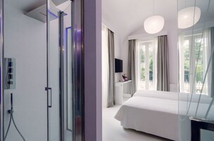 Quarto de hotel moderno com cama grande, chuveiro em cabine de vidro, TV e varanda com cortinas.