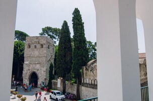 Um edifício histórico de pedra com entrada em arco, rodeado por árvores altas e um carro branco estacionado perto.