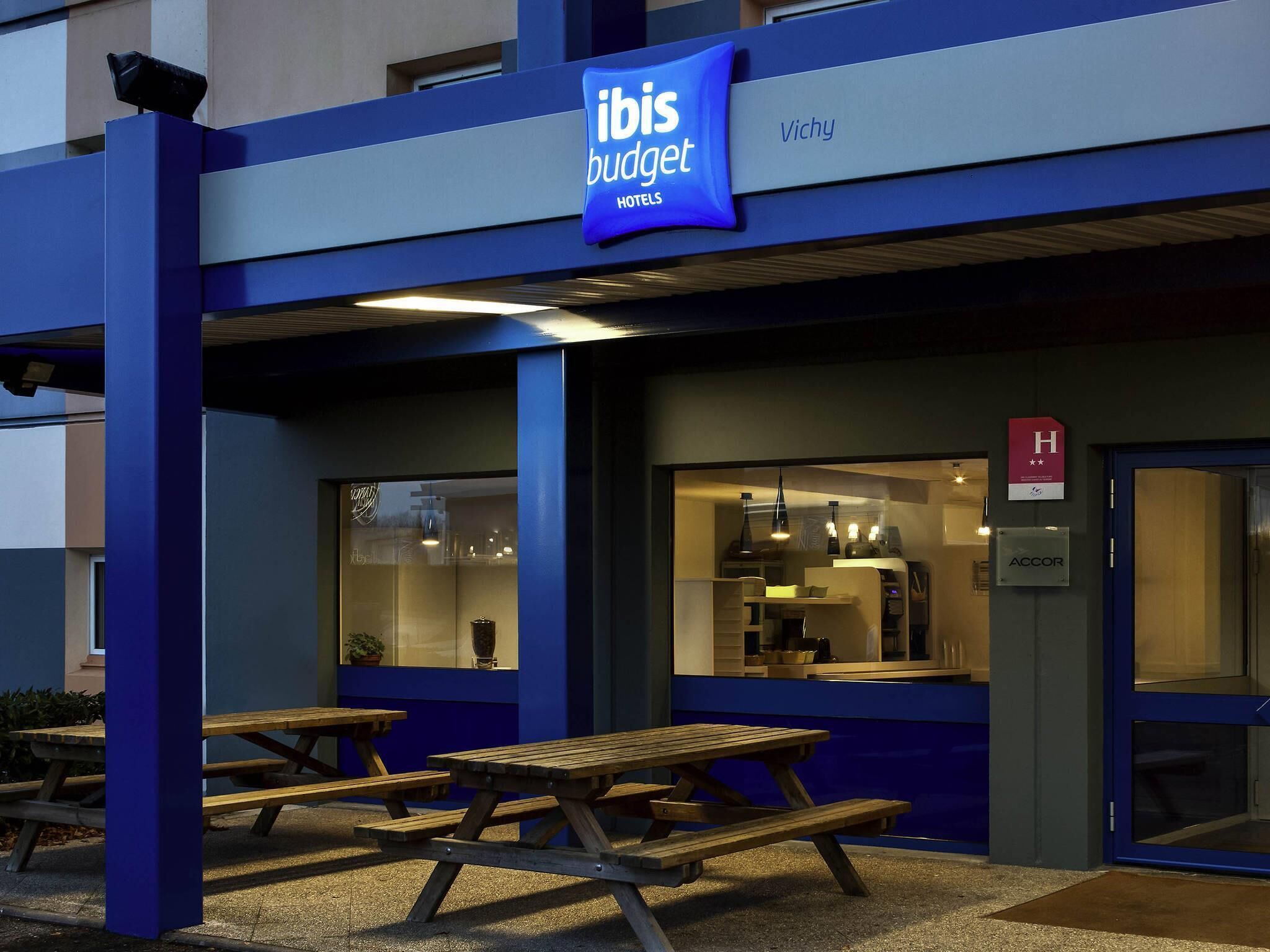 Exterior de um hotel com uma placa indicando 'ibis budget hotels' e 'Vichy'. Há áreas de estar ao ar livre com mesas e bancos de madeira.