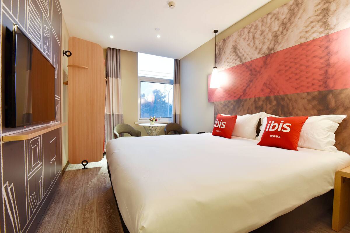 Quarto de hotel com uma cama grande, televisão, secretária e janela com cortinas.