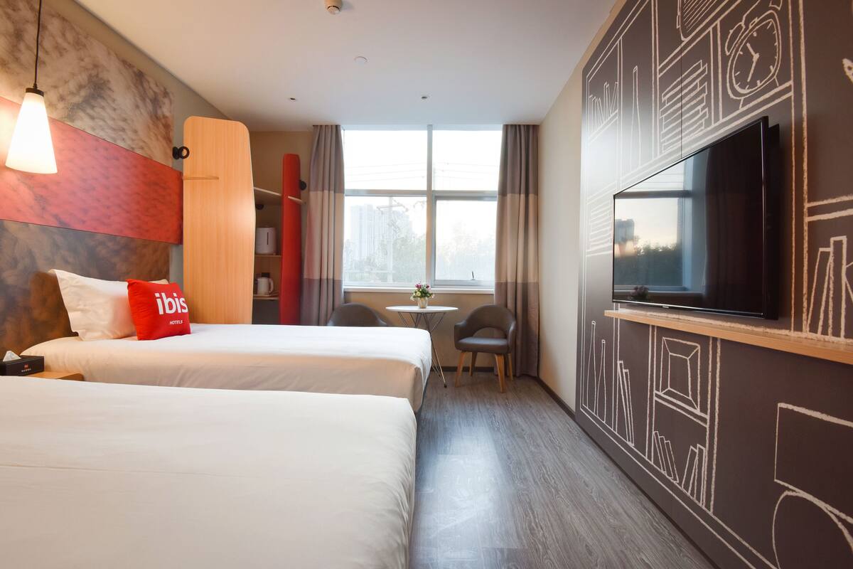 Quarto de hotel com duas camas, televisão, uma pequena mesa com cadeiras e uma janela com cortinas.