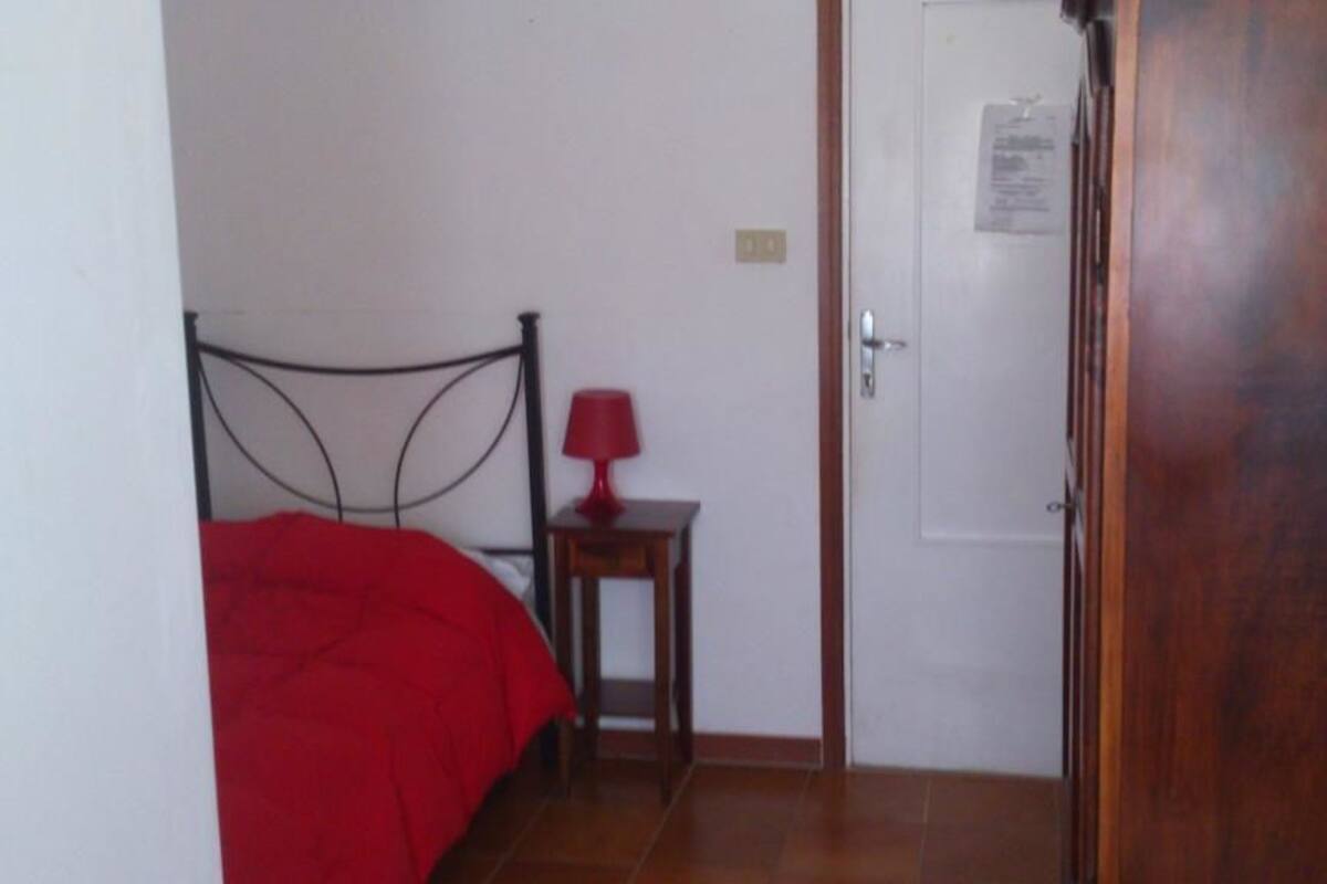 Quarto com uma cama, mesa de cabeceira, abajur vermelho e porta de madeira.