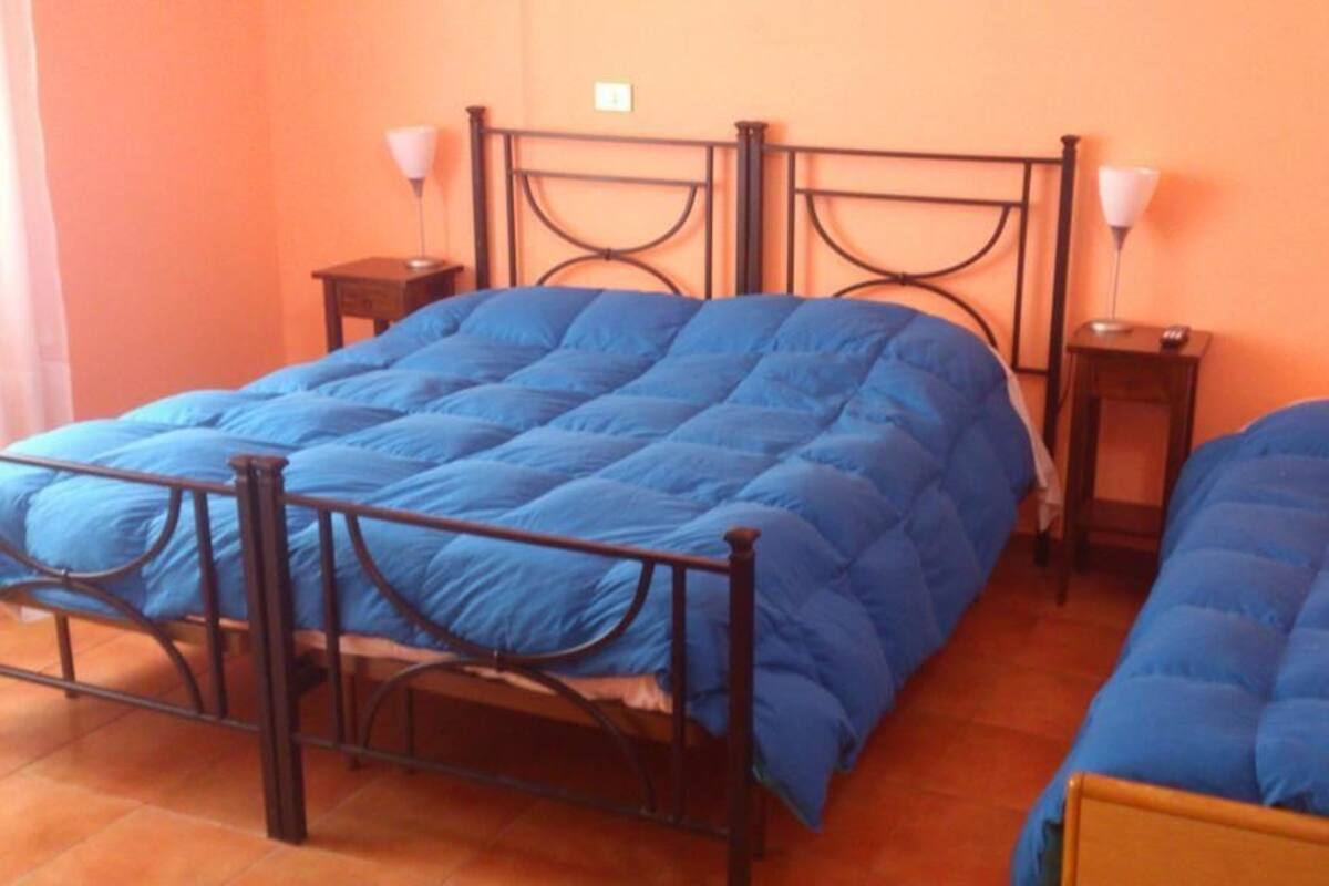 Quarto com duas camas, cada uma com colcha azul, cabeceira de madeira e uma pequena mesa de cabeceira com luminária.