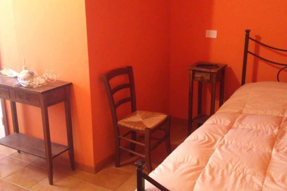 Quarto com cama, cadeira, mesa pequena e mesa de cabeceira.
